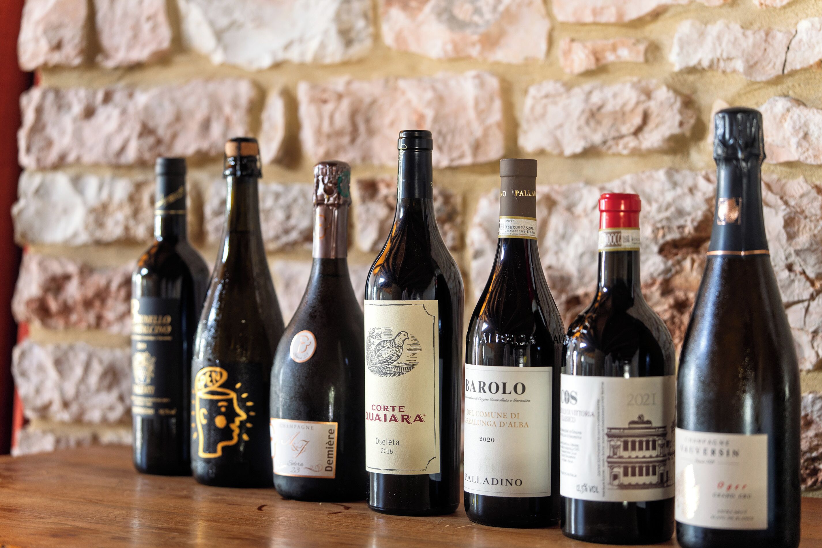 Várias garrafas de vinho dispostas numa superfície de madeira, com um muro de pedra ao fundo.