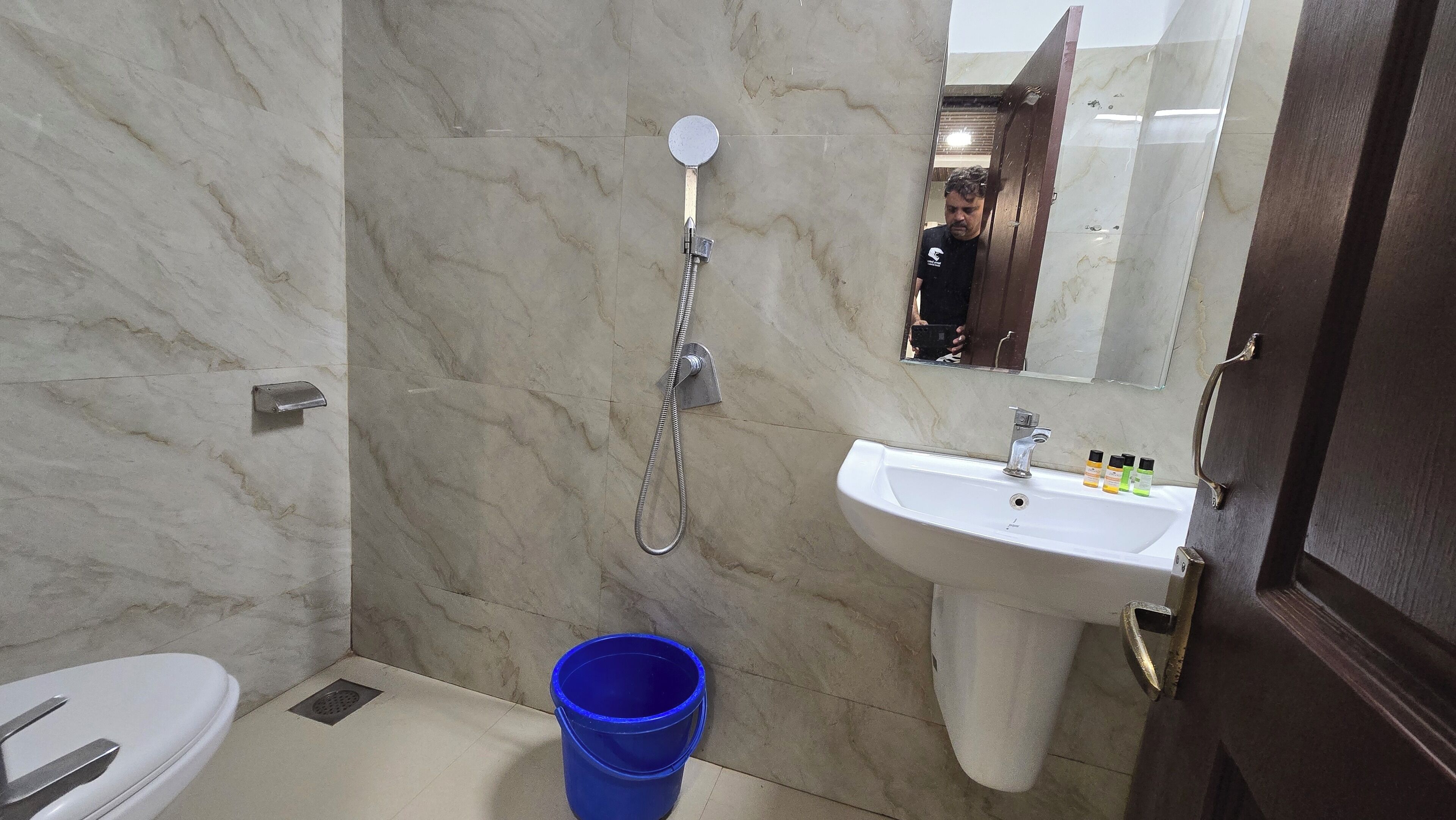 Casa de banho com lavatório branco, chuveiro manual, balde azul e sanita.