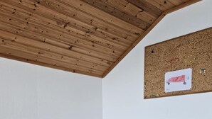 Quarto pequeno e modestamente mobiliado, com teto de madeira, uma beliche, um criado-mudo verde e um mural na parede.