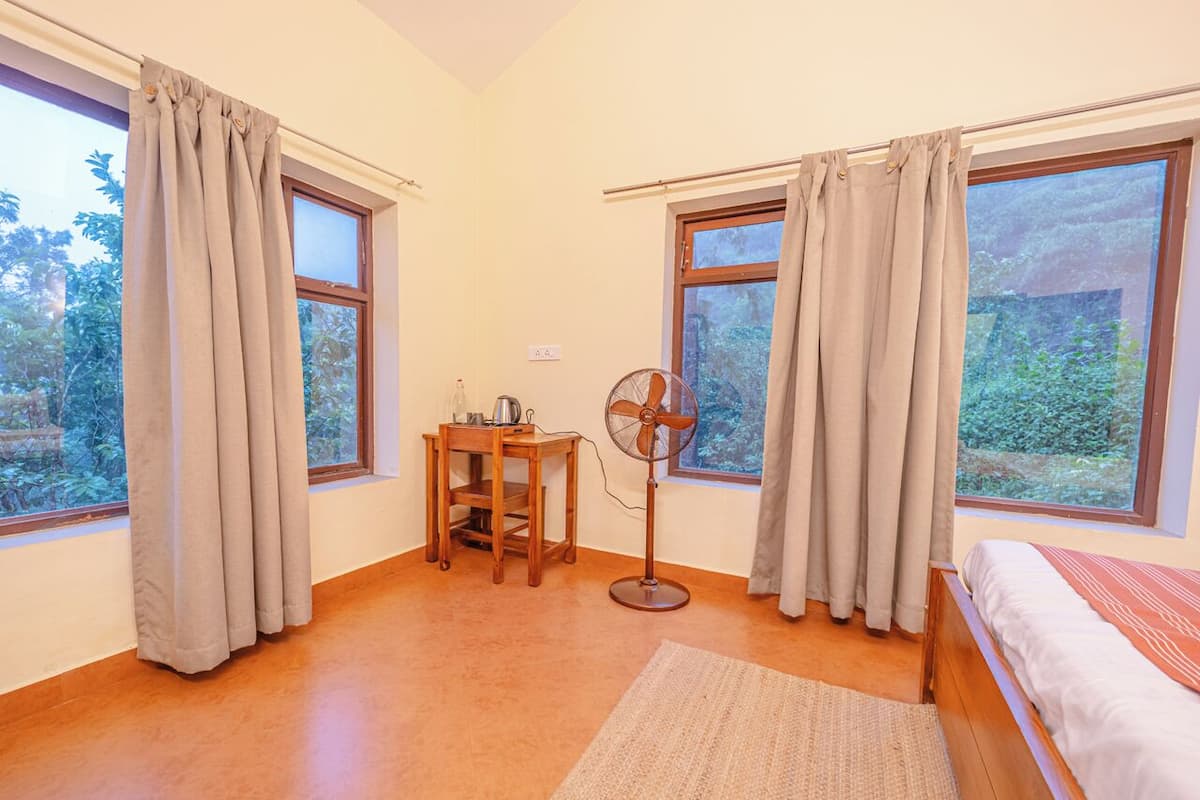 Quarto com duas janelas amplas, uma mesa de madeira com chaleira, um ventilador e uma cama com cobertor vermelho.