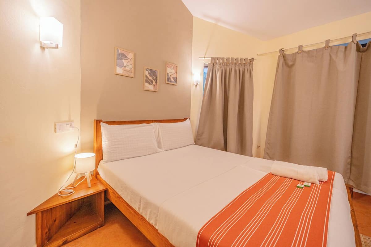 Quarto de hotel com cama, mesa de cabeceira, abajur e cortinas.