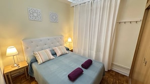 Quarto com cama, duas mesas de cabeceira, guarda-roupa e janela com cortinas.