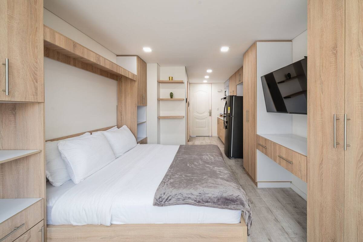Quarto compacto com cama, mesas de cabeceira, televisão e cozinha compacta.