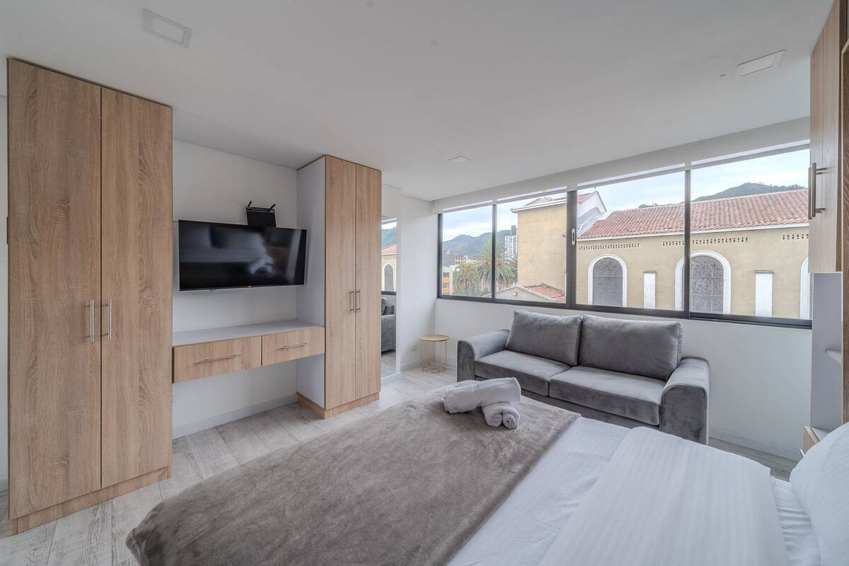 Quarto moderno com uma cama grande, um sofá cinza, um guarda-roupa de madeira, uma TV de tela plana e vista para edifícios através de grandes janelas.