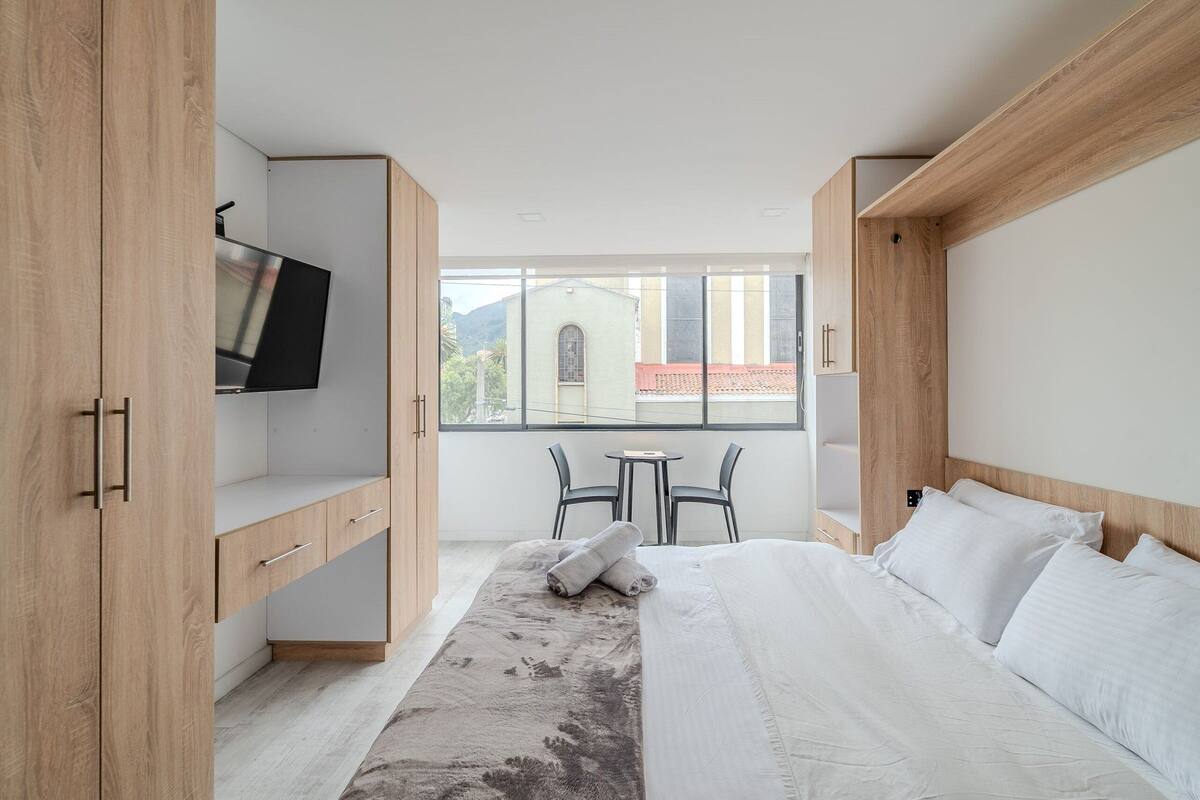 Quarto de hotel com cama, televisão, secretária e janela com vista para um edifício e montanhas.