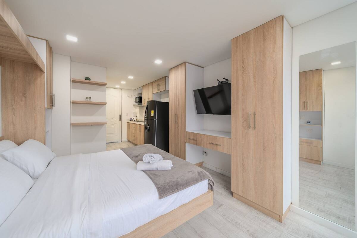Quarto moderno com cama, televisão e cozinha compacta.