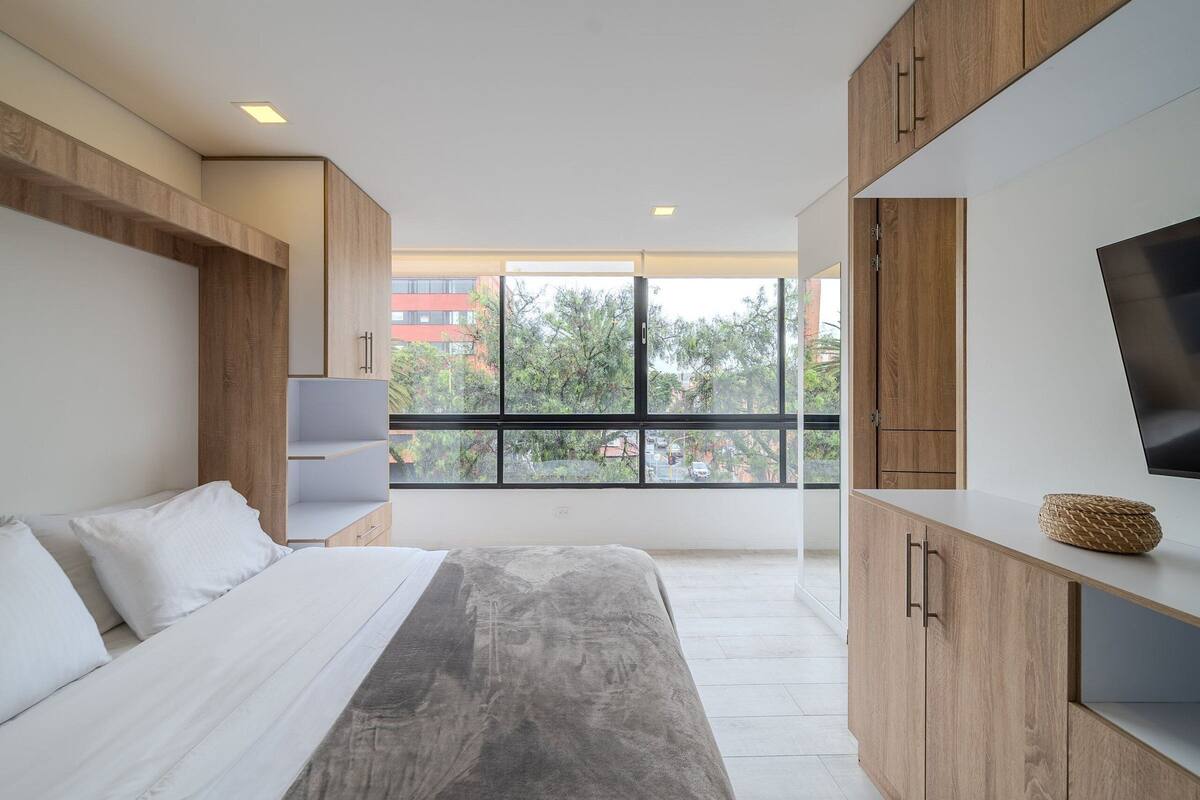 Quarto moderno com cama grande, armários embutidos de madeira, televisão de tela plana e vista da cidade através de grandes janelas.