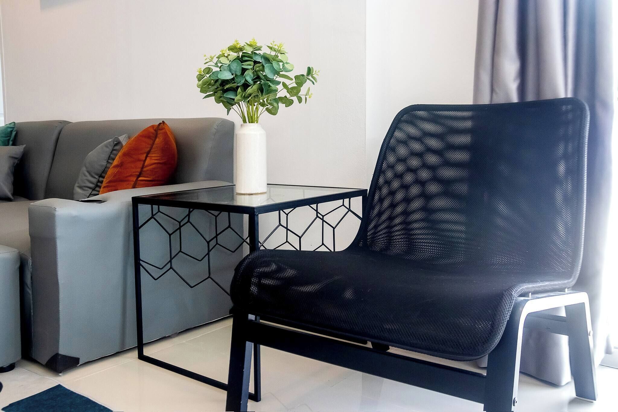Uma sala de estar moderna com um sofá cinzento, uma cadeira de malha preta, um vaso branco com uma planta e uma mesa de centro de vidro com padrão geométrico.