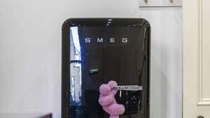 Uma geladeira preta com a marca "SMEG", localizada em uma cozinha com armários brancos e uma porta branca ao fundo.