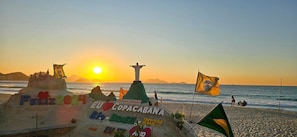 Cena de praia com um castelo de areia com a inscrição 'Feliz 2024' e 'EU COPACABANA'.
