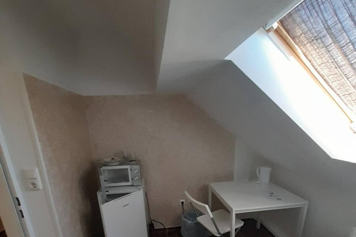Um quarto pequeno e aconchegante com teto inclinado, uma cama, uma escrivaninha, uma cadeira e um micro-ondas.