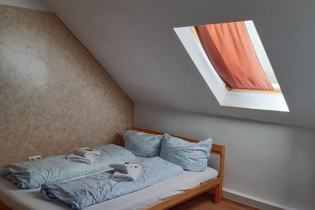 Quarto com teto inclinado, cama com estrutura de madeira e janela com cortina.