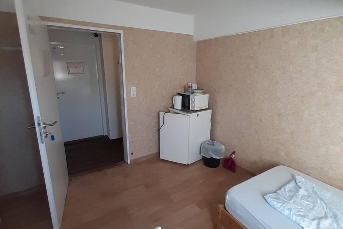 Quarto pequeno e pouco mobiliado, com piso de madeira, uma cama, uma escrivaninha com cadeira, um frigorífico branco pequeno e uma porta que dá para outro cômodo.