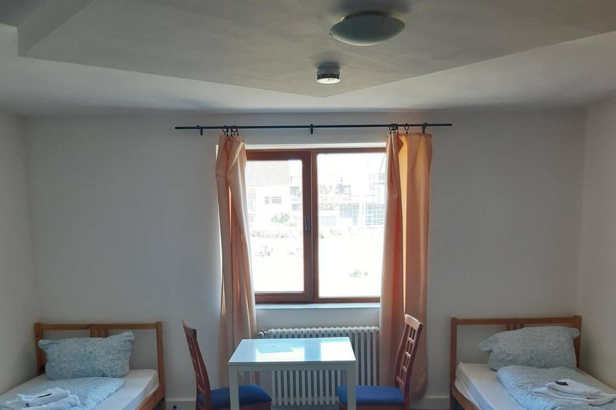 Quarto com duas camas de solteiro, uma mesa com cadeiras, um radiador e uma janela com cortinas.