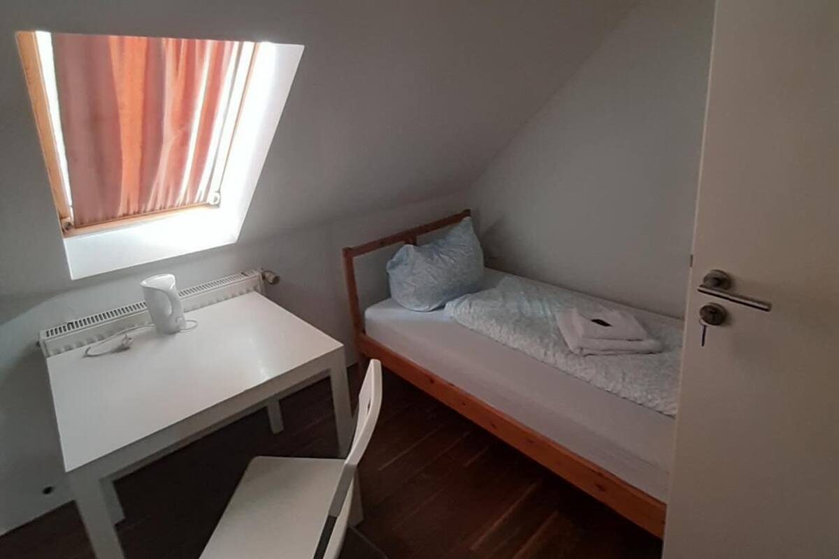 Quarto pequeno e funcional, com uma cama de solteiro, escrivaninha com cadeira, radiador e janela com cortinas.