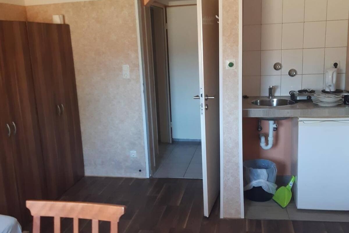 Quarto compacto com cama, uma mesinha, cozinha compacta com pia e fogão, e uma porta que dá para outro cômodo.