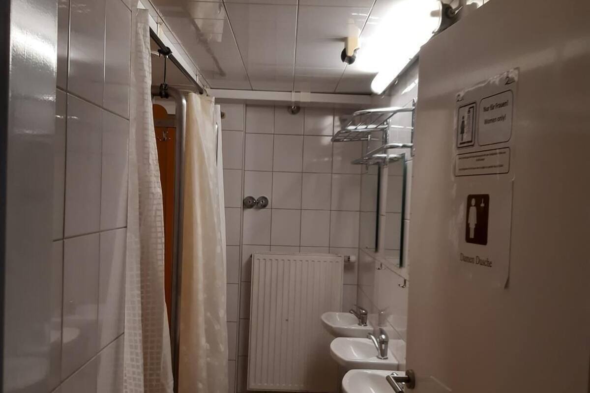 Casa de banho pequena com paredes e chão em azulejo branco, cortina de chuveiro, radiador e duas bacias com espelhos.