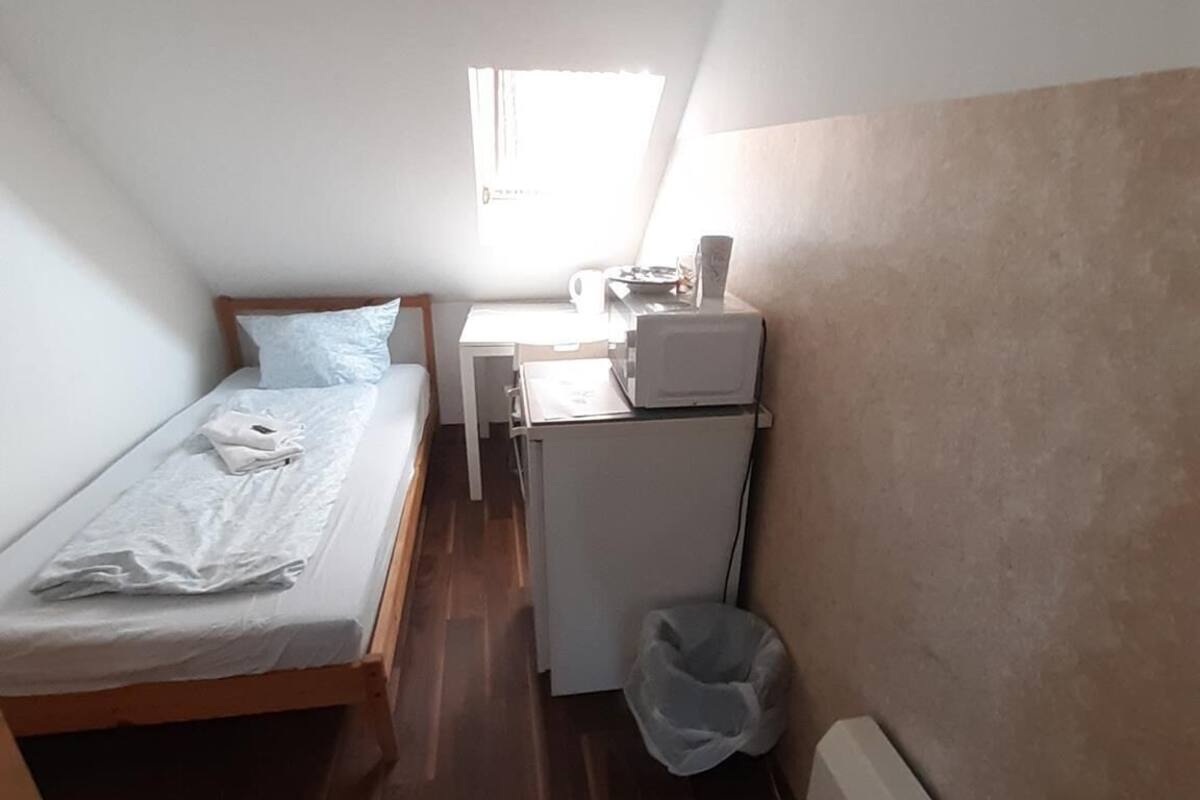 Quarto pequeno e estreito com uma cama, uma escrivaninha, um micro-ondas e uma lixeira.