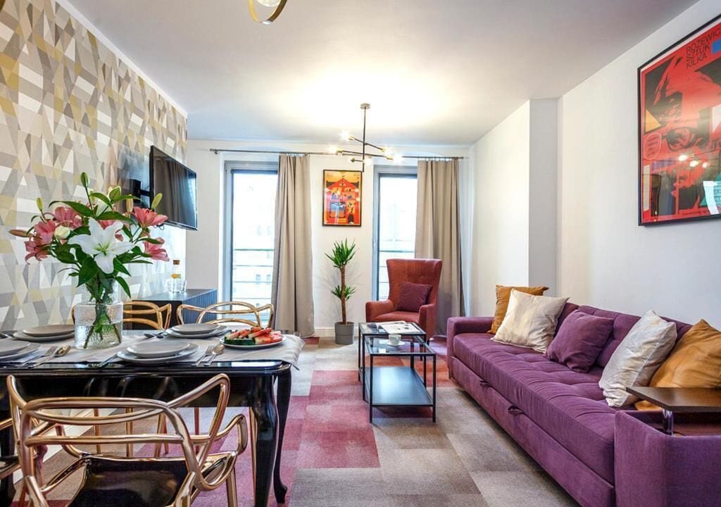 Uma sala de estar moderna com um sofá roxo, uma mesa de jantar para quatro pessoas e uma TV fixada na parede.