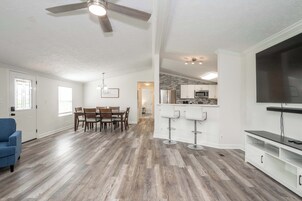 Ampla sala de estar com uma mesa de jantar, cadeiras e um sofá azul. Uma cozinha com micro-ondas e banquetas pode ser vista ao fundo.