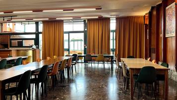Uma sala de jantar espaçosa com várias mesas e cadeiras, grandes janelas com cortinas, e uma cozinha visível ao fundo.