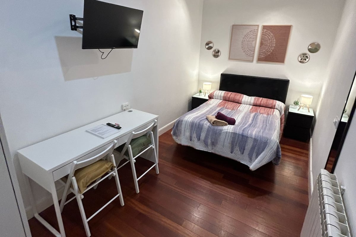 Quarto de hotel pequeno com cama, escrivaninha, televisão e radiador.