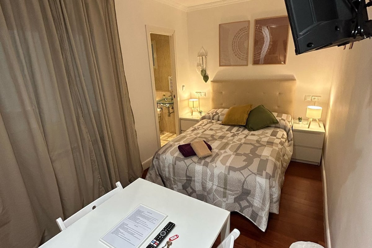 Quarto de hotel com cama, mesa de cabeceira, cadeira, televisão e um pequeno cesto de lixo.