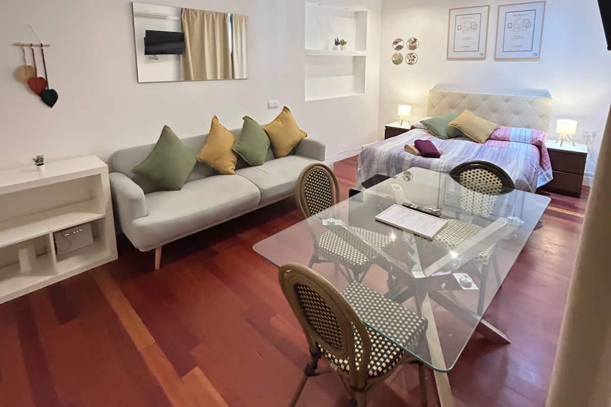 Uma sala de estar moderna com uma mesa de jantar de vidro, um sofá com almofadas, uma TV fixada na parede e uma luminária no teto.