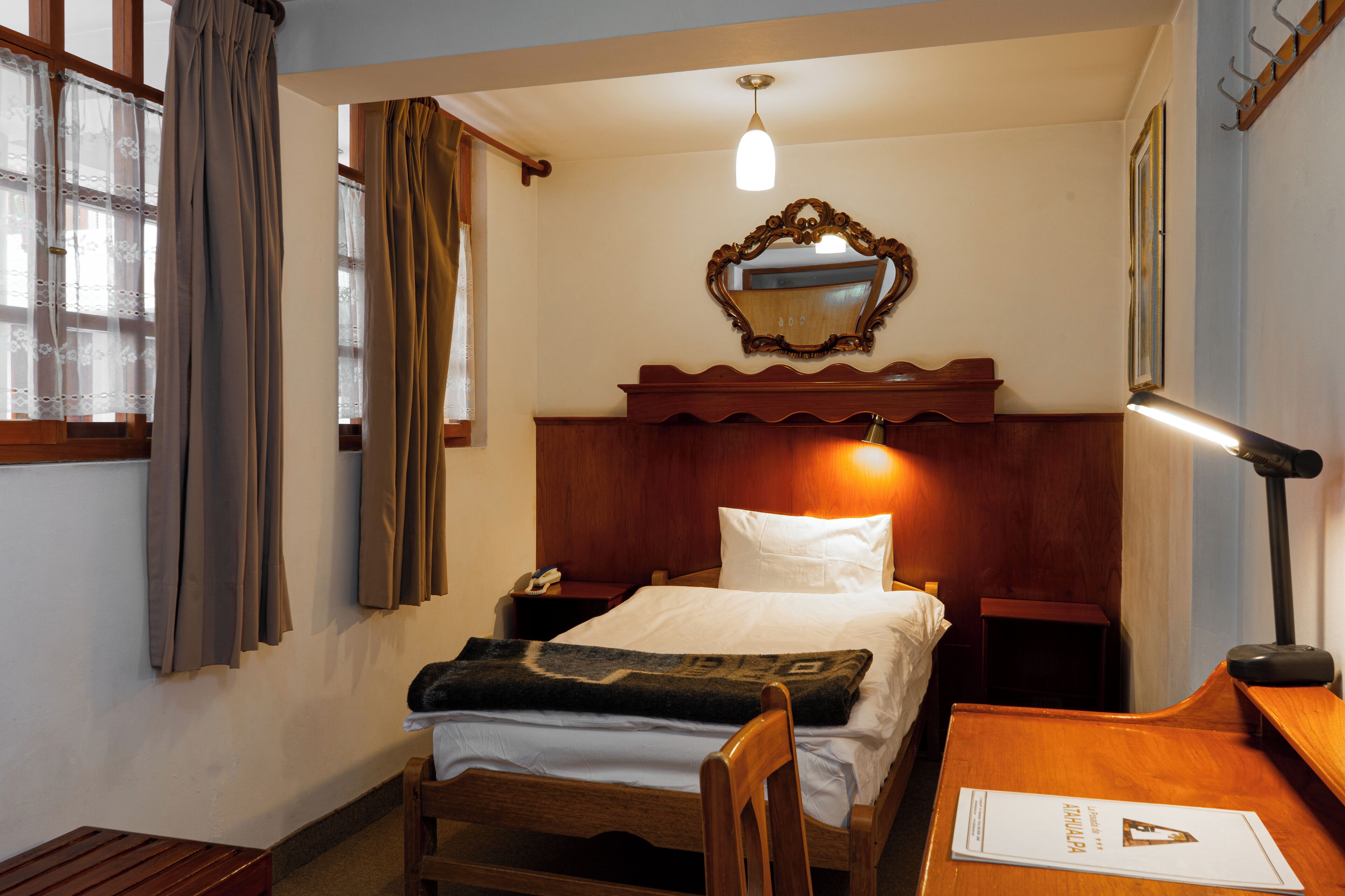 Quarto de hotel com cama de madeira, mesa de cabeceira com abajur, criado-mudo com telefone e espelho fixado na parede.