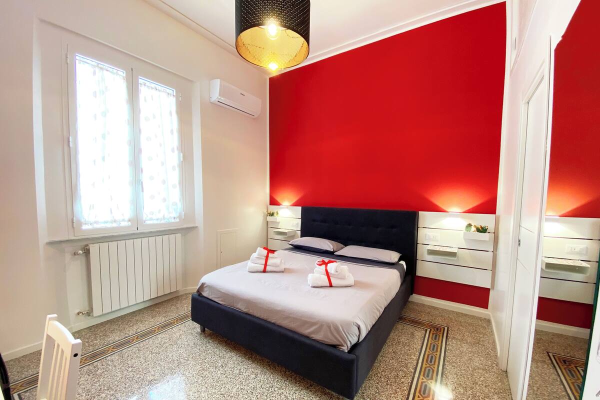 Quarto com cama, mesas de cabeceira brancas, uma parede de destaque vermelha e uma janela com cortinas.