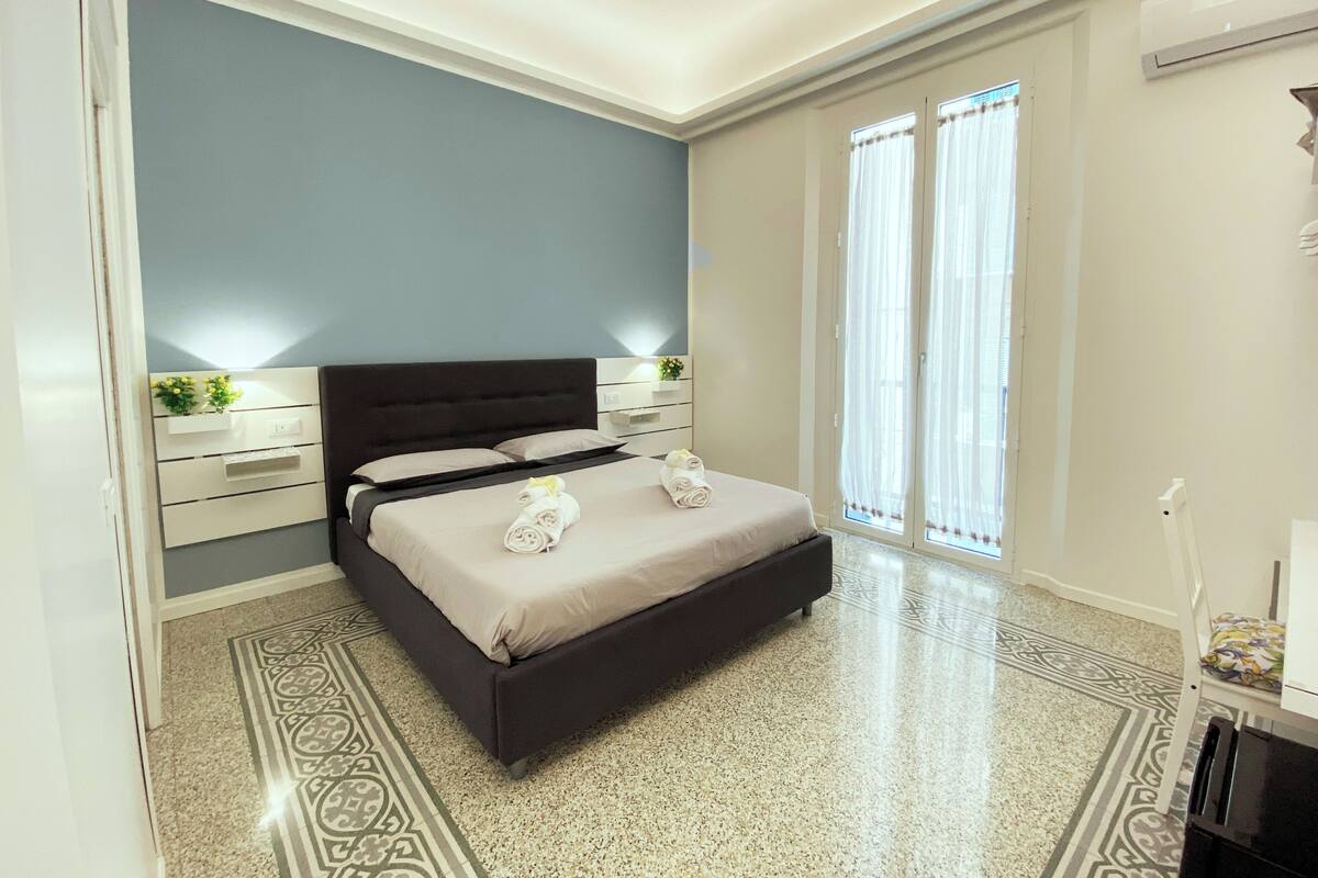 Quarto com cama, mesas de cabeceira, rodapé, janela com cortinas e piso estampado.