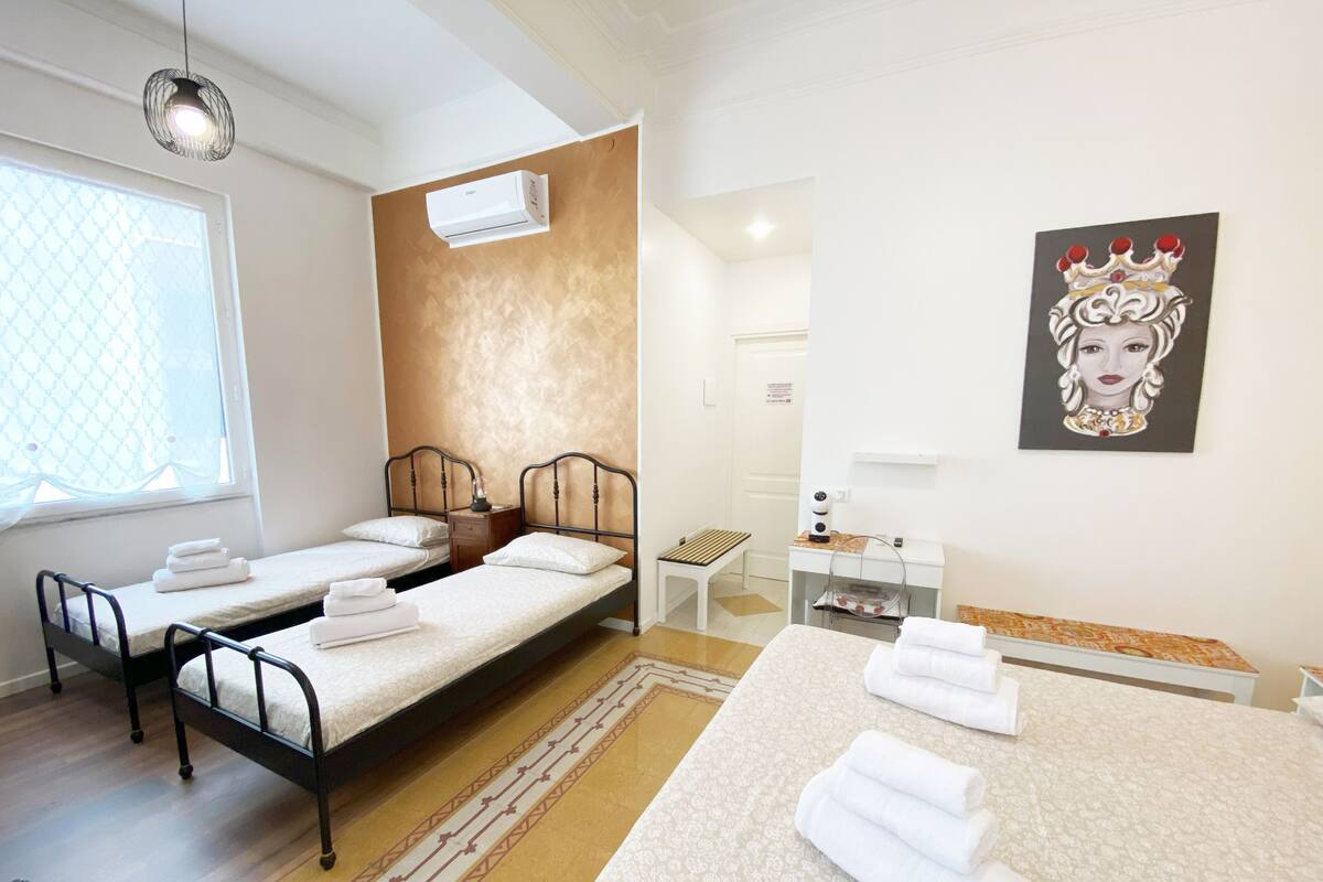 Quarto de hotel com duas camas de solteiro, uma janela grande com vista, ventilador de teto, ar condicionado, mesa de cabeceira com abajur, pequena mesa de cabeceira com objeto decorativo e obra de arte na parede.