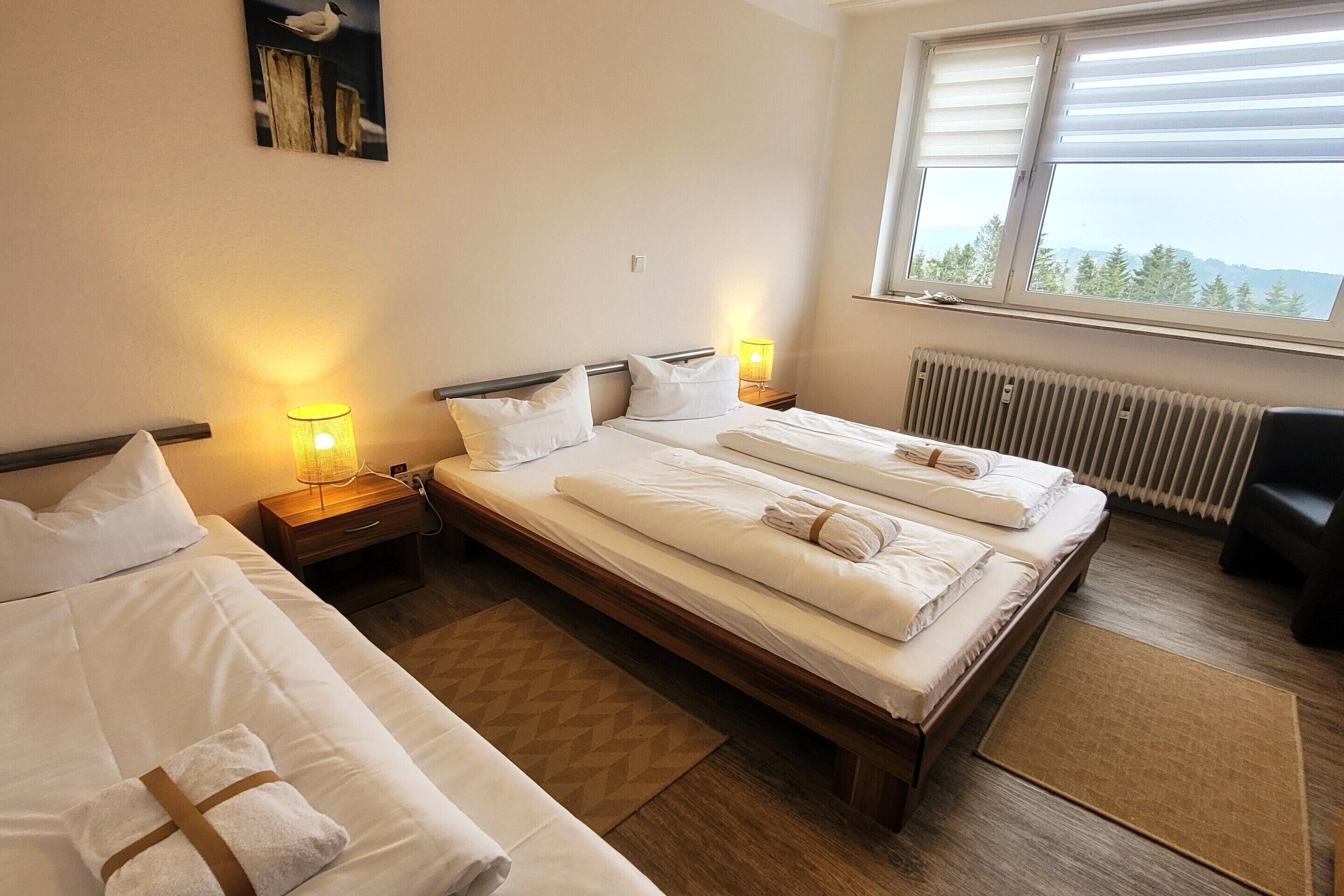 Quarto de hotel com duas camas de solteiro, piso de madeira, radiador e janela com vista para árvores.