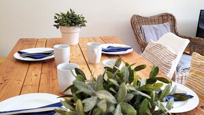 Uma mesa de madeira posta com pratos, talheres e copos brancos, com um vaso de planta em primeiro plano.