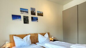 Quarto de hotel com duas camas, cabeceira de madeira, roupa de cama branca, mesa de cabeceira com abajur e seis quadros na parede.