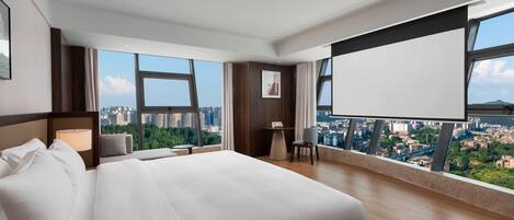 Quarto de hotel moderno com uma cama grande, uma mesa de trabalho e vista para o horizonte urbano através de amplas janelas.