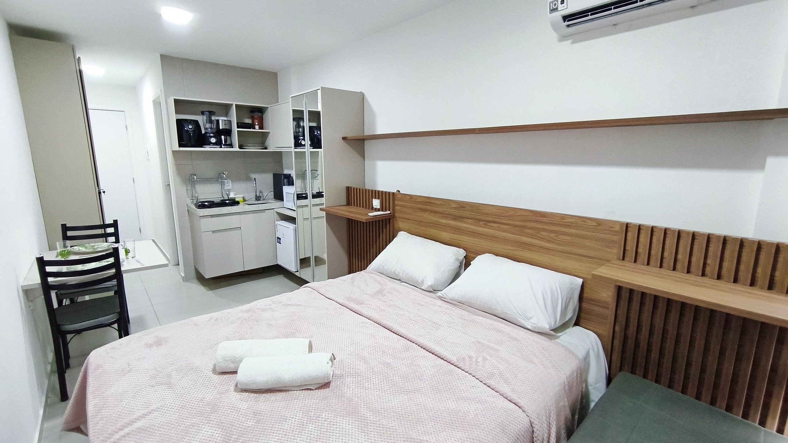 Quarto com cama, mesa de cabeceira e área de cozinha compacta, incluindo micro-ondas, cafeteira e armários.