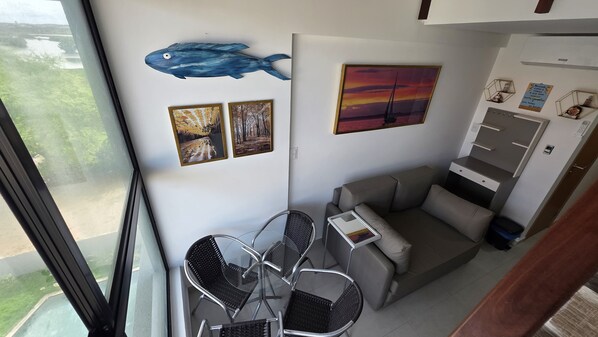 Quarto com mesa e cadeiras de vidro, um sofá cinza, uma pintura do pôr do sol e um enfeite de peixe azul na parede.