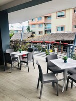 Um pátio externo com mesas e cadeiras, uma estrutura com telhado de palha e vista para edifícios de hotel ao fundo.