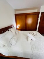 Quarto de hotel com uma cama grande, cabeceira de madeira, mesa de cabeceira com abajur e guarda-roupa.