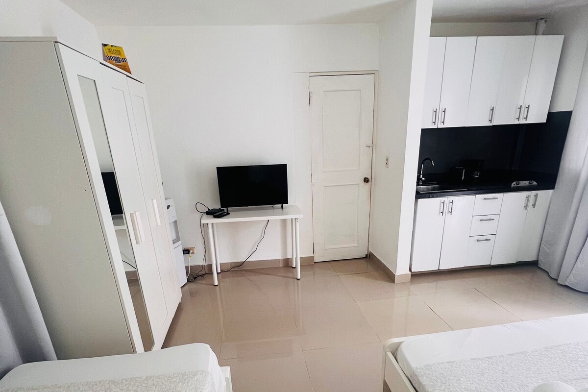 Quarto de hotel com duas camas, televisão, escrivaninha e cozinha compacta.