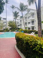 Uma área de piscina com espreguiçadeiras, palmeiras e um edifício de vários andares com varandas.