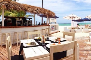 Restaurante à beira-mar com mesas e cadeiras de madeira, área de bar e guarda-sóis para proteção do sol.