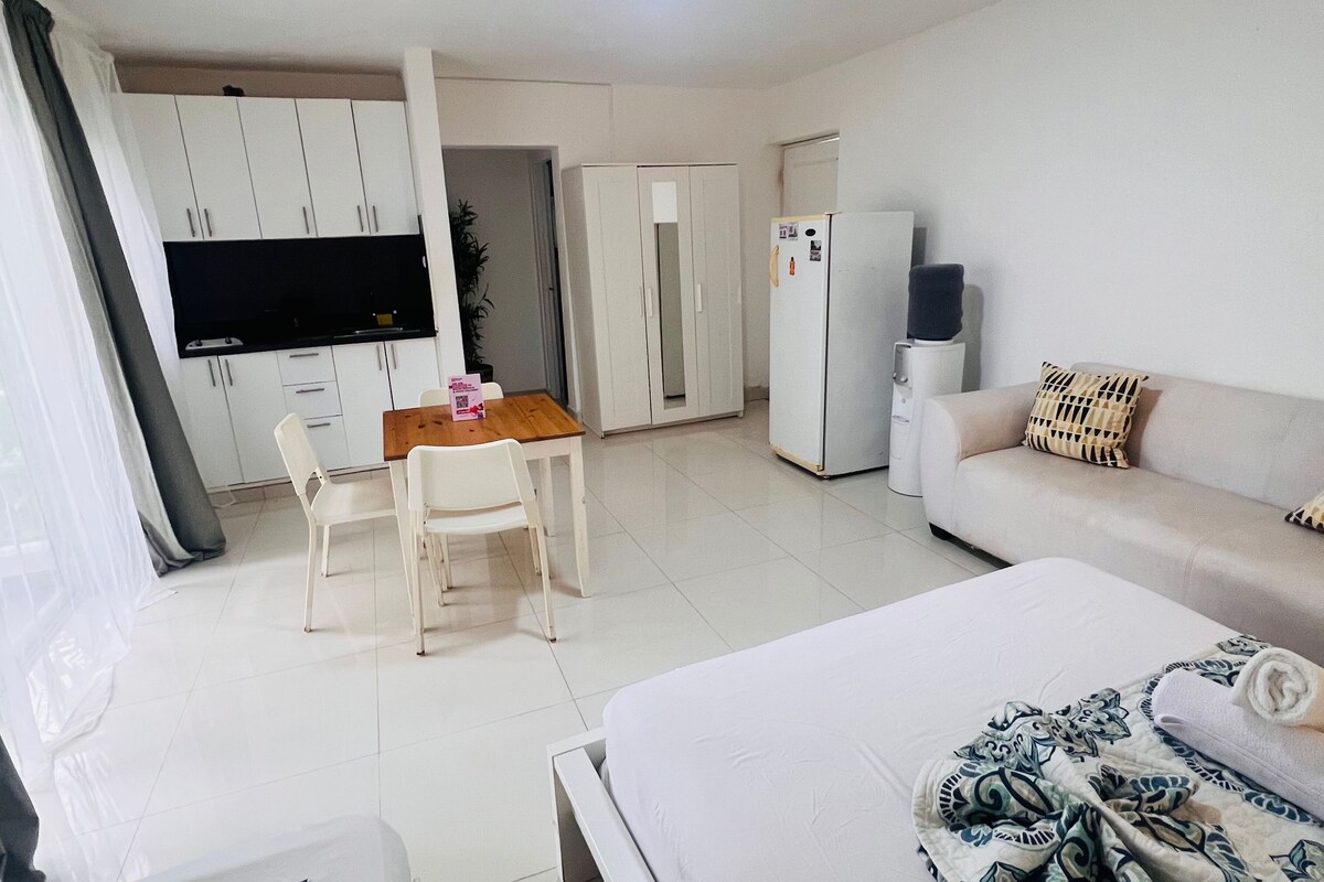 Quarto de hotel com cama, sofá, mesa de jantar com cadeiras, televisão e cozinha compacta.
