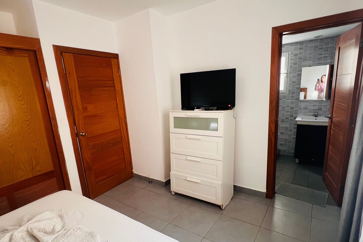 Quarto com cama, cômoda, televisão e banheiro visível por uma porta aberta.