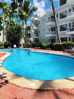 Piscina com espreguiçadeiras e palmeiras, rodeada por um edifício com várias andares e sacadas.