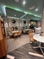 Uma sala de jantar com mesas redondas e cadeiras de vime, uma mesa de recepção e uma planta ao fundo.