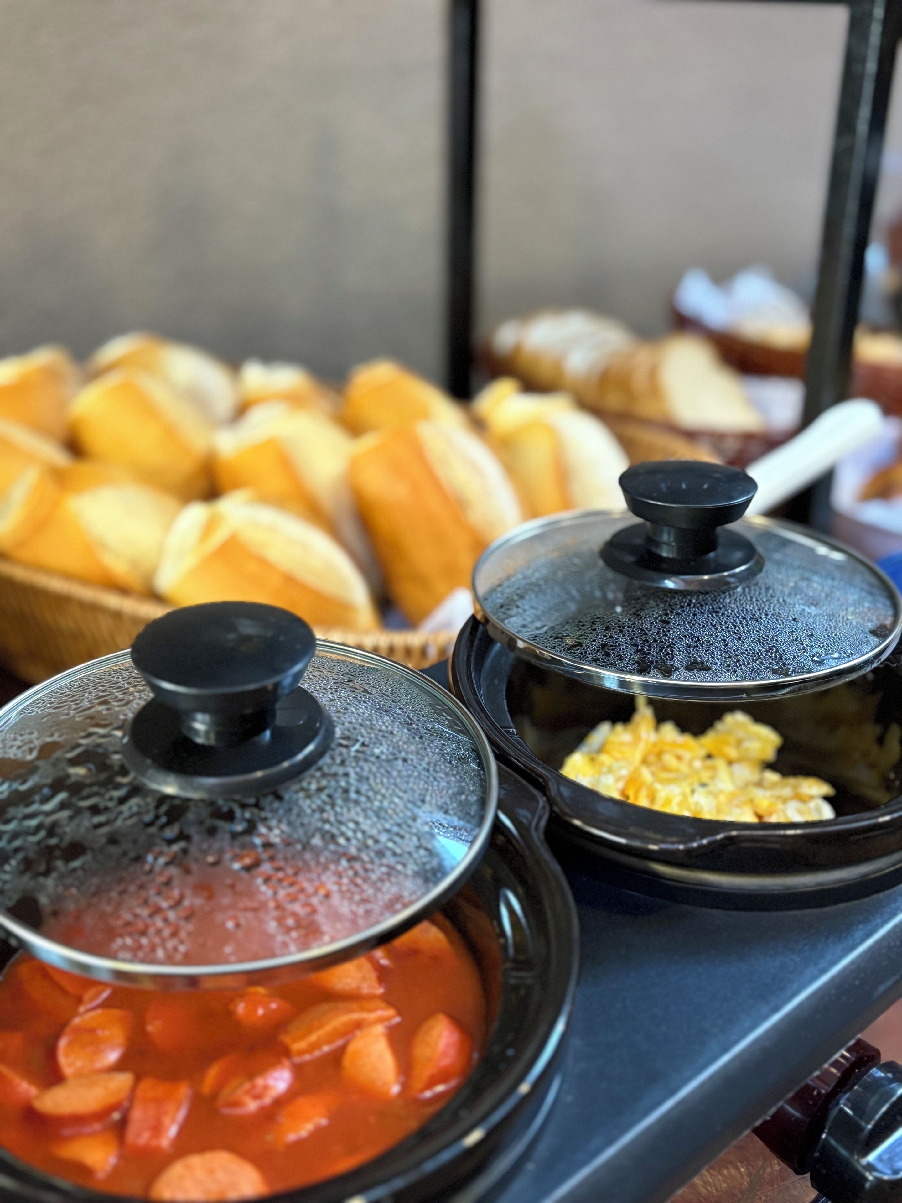 Disposição de buffet com diversos alimentos, incluindo uma panela de sopa, um recipiente com ovos mexidos e uma cesta de pães.