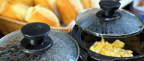 Disposição de buffet com diversos alimentos, incluindo uma panela de sopa, um recipiente com ovos mexidos e uma cesta de pães.