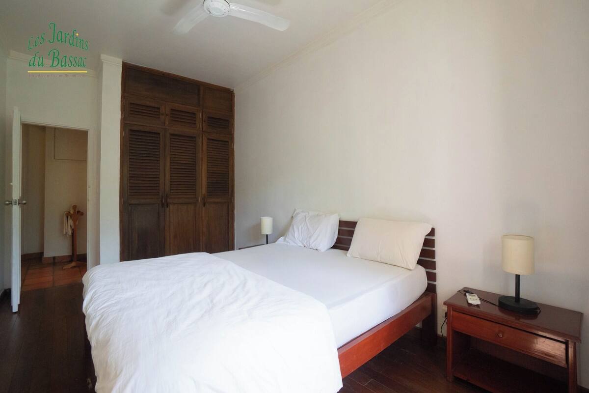 Quarto com cama grande, guarda-roupa de madeira, mesa de cabeceira e ventoinha no teto.